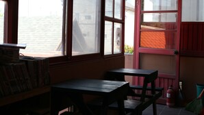 Quarto com mesa e cadeiras de madeira, estante, janela com vista para um edifício e porta vermelha.