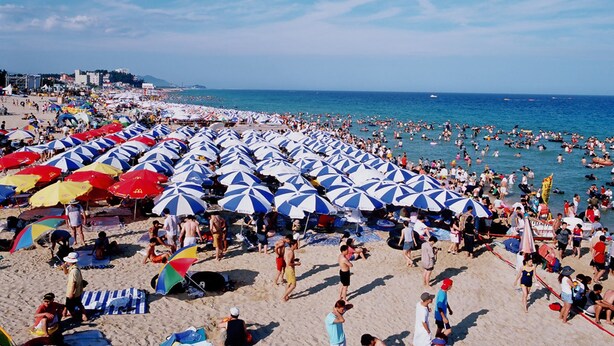 Uma praia cheia, com vários guarda-sóis e pessoas aproveitando a água.