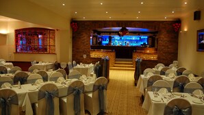 Uma sala de banquetes com mesas preparadas para um evento formal, área de bar com parede de tijolos e uma janela decorativa com luzes vermelhas.