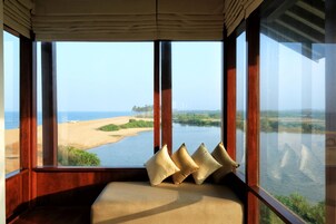 Quarto com uma grande janela com vista para uma praia e um rio. Há quatro almofadas em um banco.