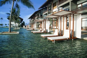Um resort com espreguiçadeiras ao redor da piscina, um caminho ladeado por palmeiras e um edifício com varandas com vista para a água.