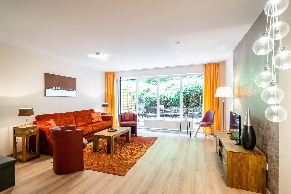 Uma sala de estar moderna com um sofá vermelho, mesa de centro de madeira e vista para o verde lá fora.
