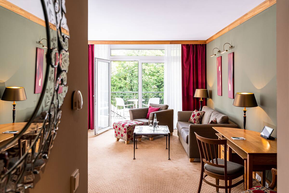 Uma sala de estar aconchegante com uma mesa de madeira, um sofá e uma poltrona. Há uma janela com cortinas, um abajur e uma peça decorativa na parede.