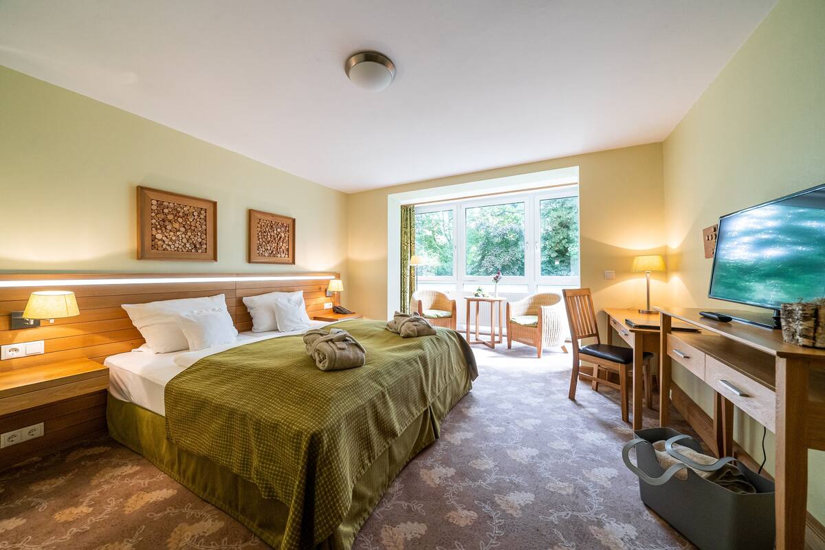 Quarto de hotel com cama, escrivaninha, cadeira, televisão e janela com vista para árvores.