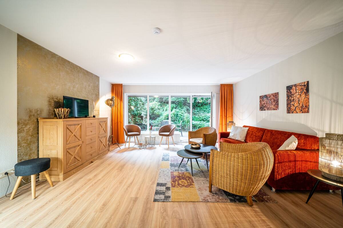 Uma sala de estar ampla, com piso de madeira, um sofá vermelho, cadeiras de vime e uma mesa de jantar.