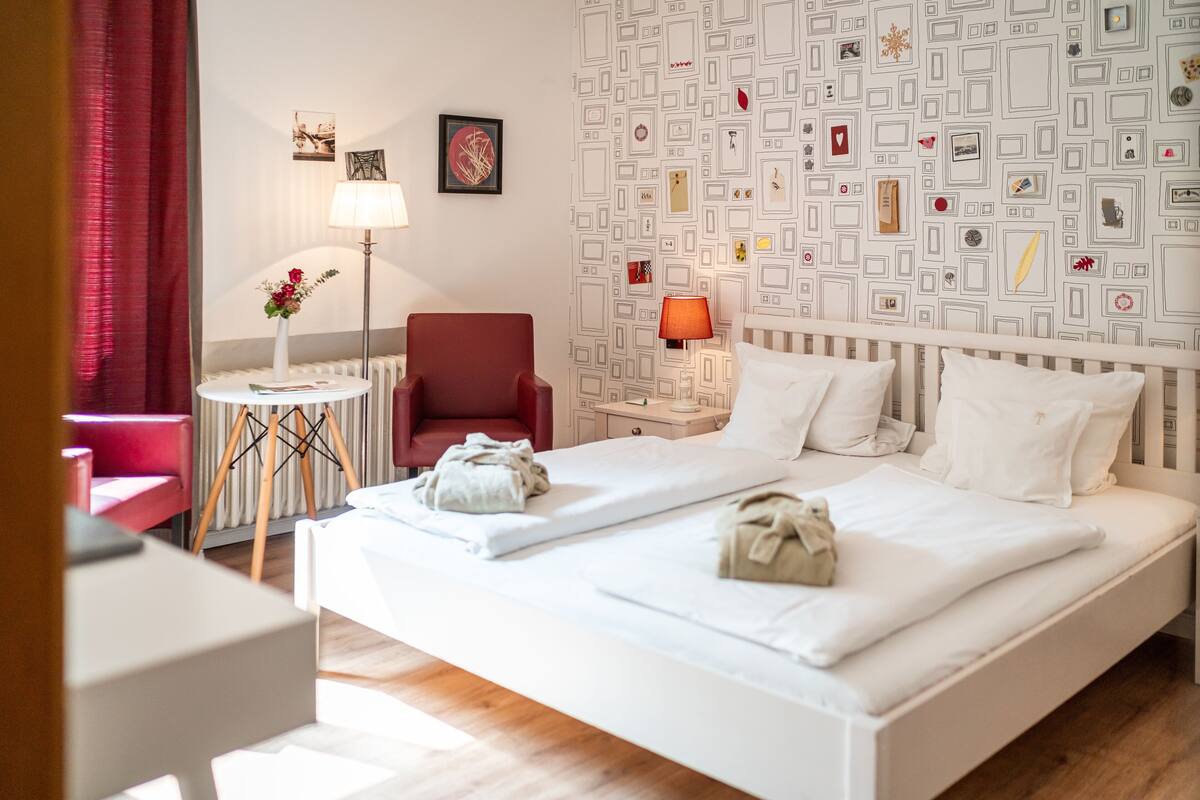 Quarto de hotel com uma cama grande, uma poltrona vermelha, uma pequena mesa redonda com abajur e uma parede com papel de parede estampado.
