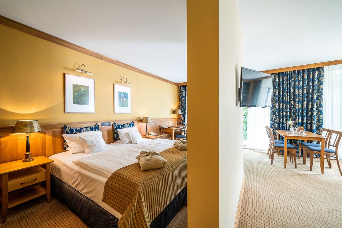Quarto de hotel com cama, mesa de cabeceira, abajur e escrivaninha com cadeira. Há uma TV na parede e uma janela com cortinas.