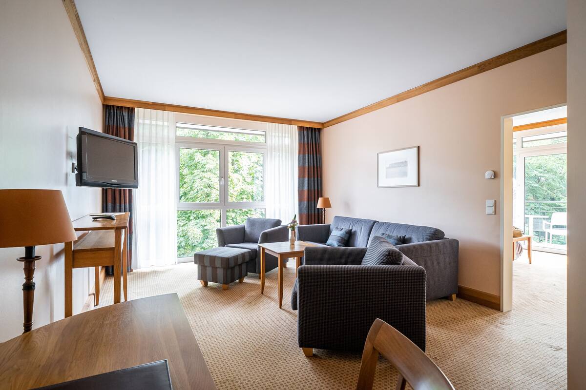 Quarto de hotel moderno com uma janela grande, TV de tela plana, sofá, poltrona e uma pequena mesa.