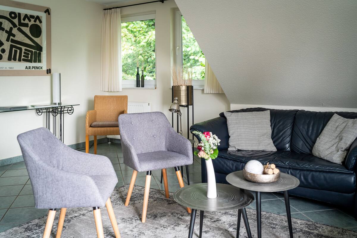 Uma sala de estar moderna com dois poltronas cinzas, um sofá de couro preto, uma mesa de centro redonda e um vaso com flores.
