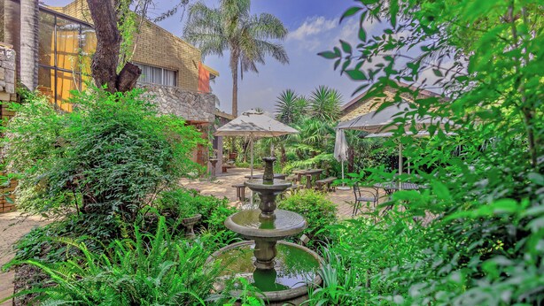 Pátio com uma fonte central, cercado por vegetação exuberante e um edifício com telhado de palha.