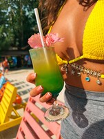 Uma pessoa segurando um drink com canudo e uma flor rosa, vestindo uma blusa amarela e um colar com pingentes.