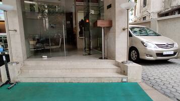 Entrada de um edifício com um carro estacionado na frente, um tapete verde e um poste de luz.