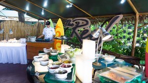 Buffet com diversos pratos, um chef e um cenário tropical.