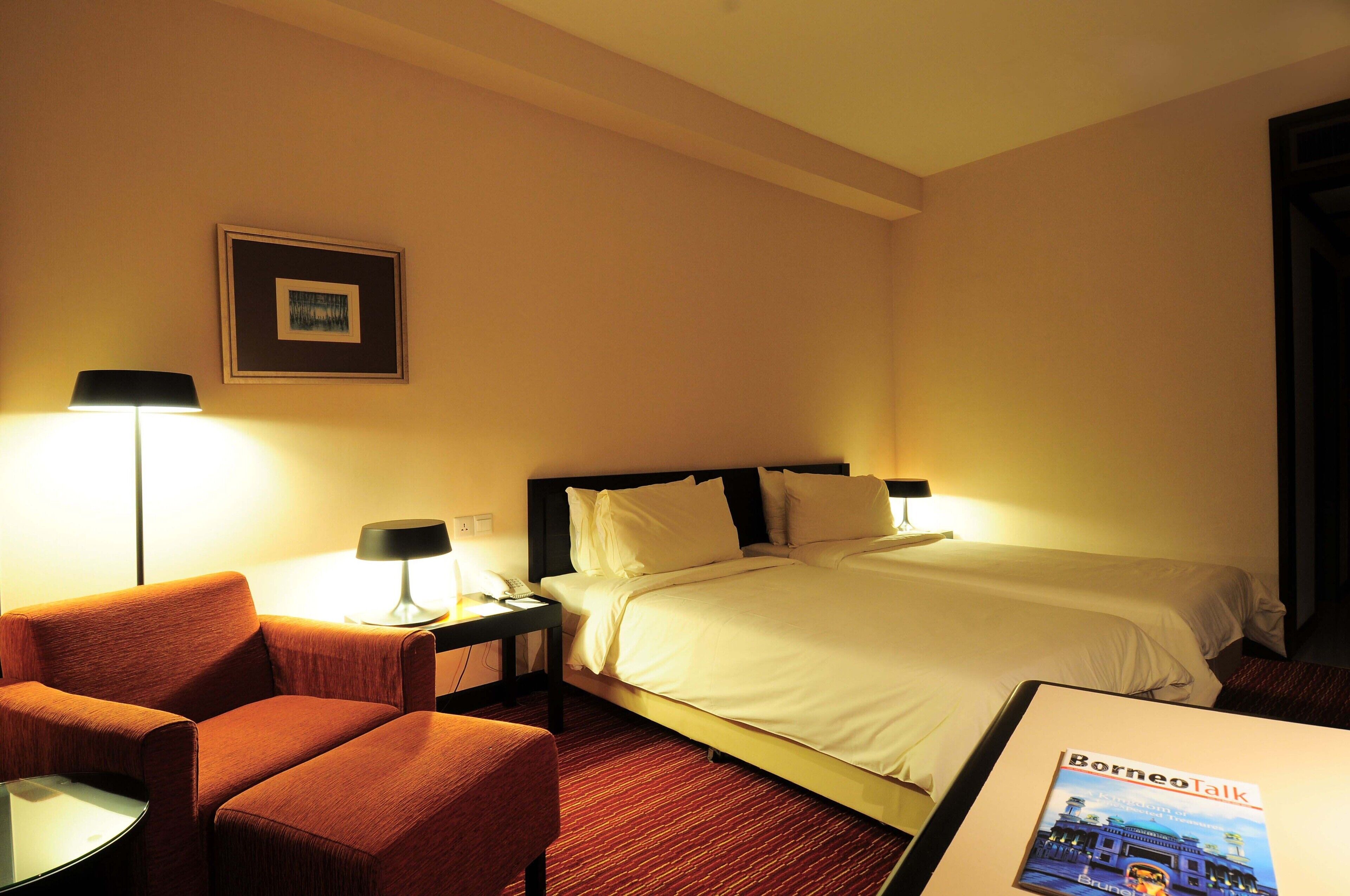 Quarto de hotel com cama, poltrona, mesa de cabeceira, abajur e um quadro na parede.