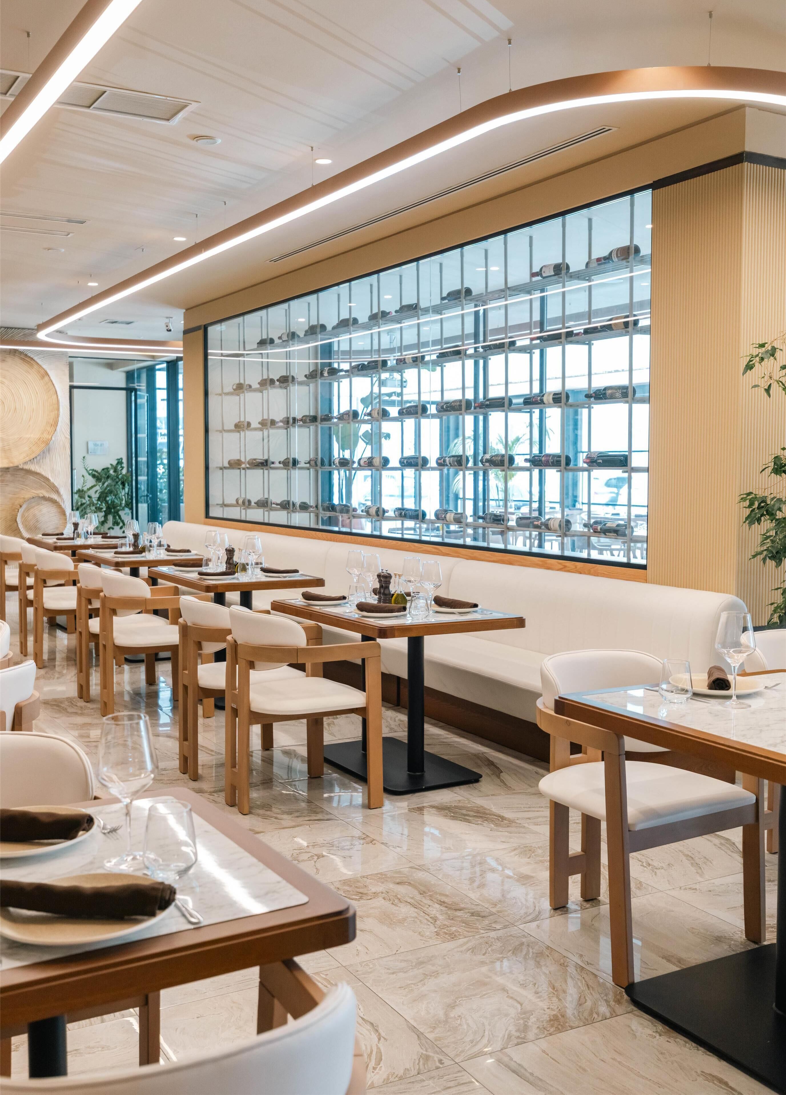 Um restaurante moderno com um balcão comprido, cadeiras brancas e uma vitrine de vidro com garrafas.
