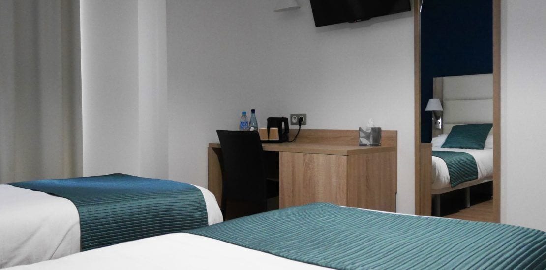Quarto de hotel com duas camas de solteiro, uma escrivaninha com cadeira, televisão de tela plana e uma mesinha com abajur e uma garrafa de água.