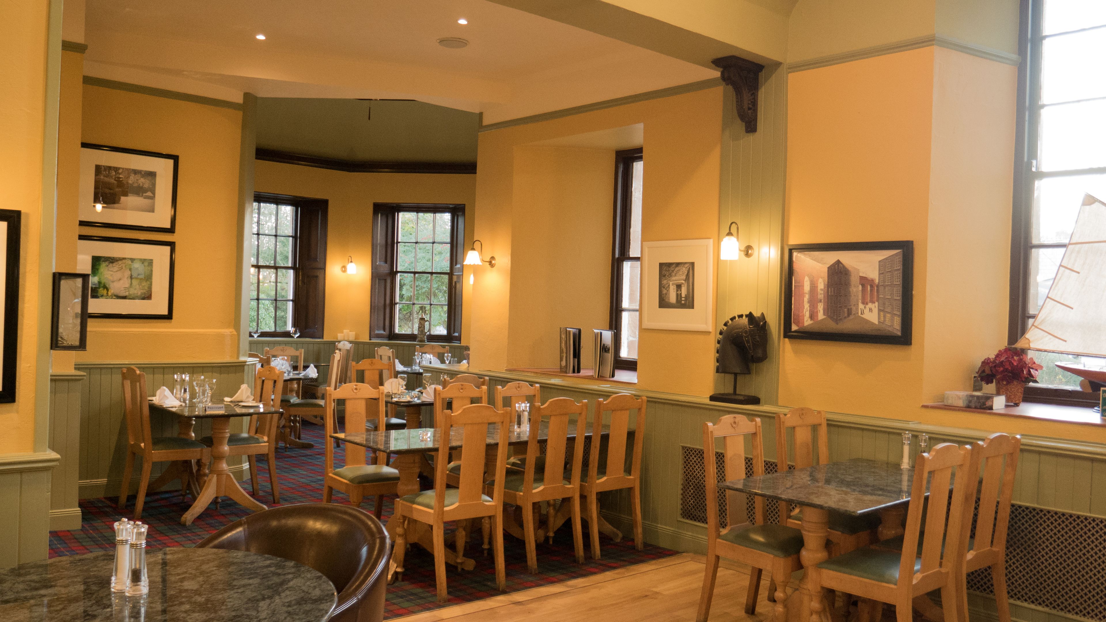 Restaurante com mesas e cadeiras de madeira, quadros nas paredes e grandes janelas que permitem a entrada de luz natural.