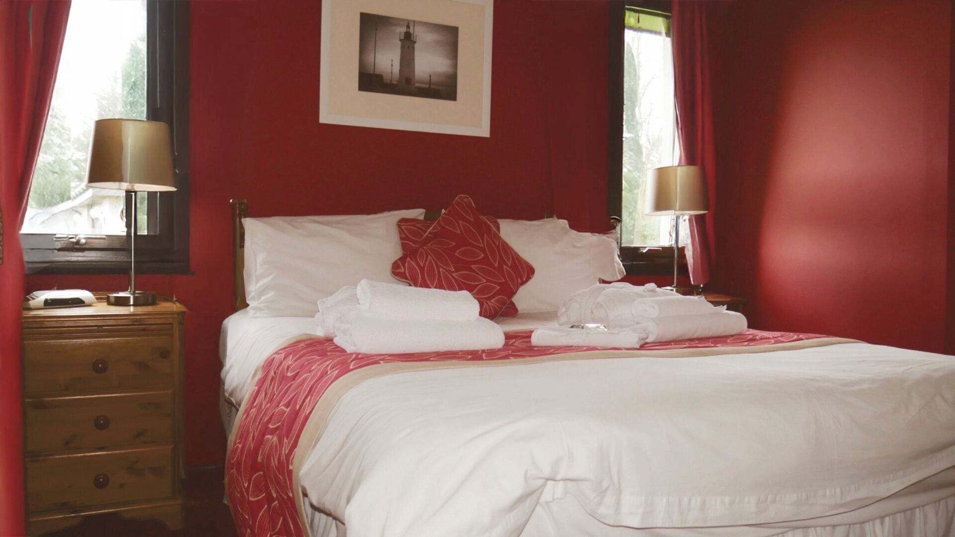 Uma cama bem arrumada com lençóis brancos e almofada estampada em vermelho, uma mesa de cabeceira de madeira com uma luminária, e um quadro na parede.