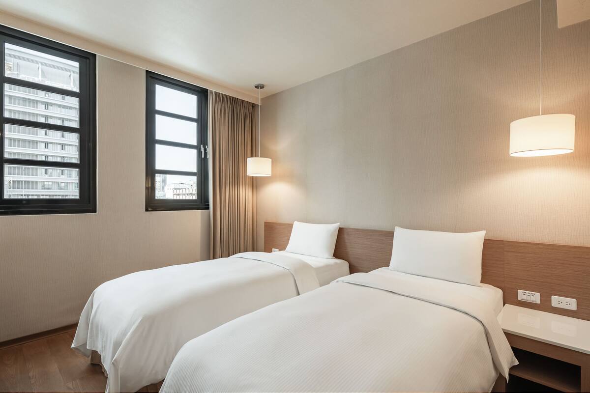 Quarto de hotel com duas camas, janela com cortinas, luminária na parede e cabeceira de madeira.