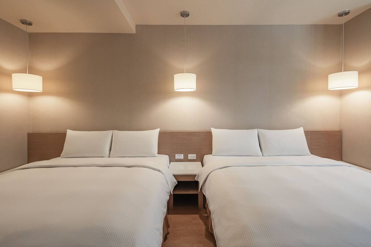 Quarto de hotel com duas camas, cada uma com uma mesa de cabeceira, uma luminária fixada na parede e uma cabeceira de madeira.