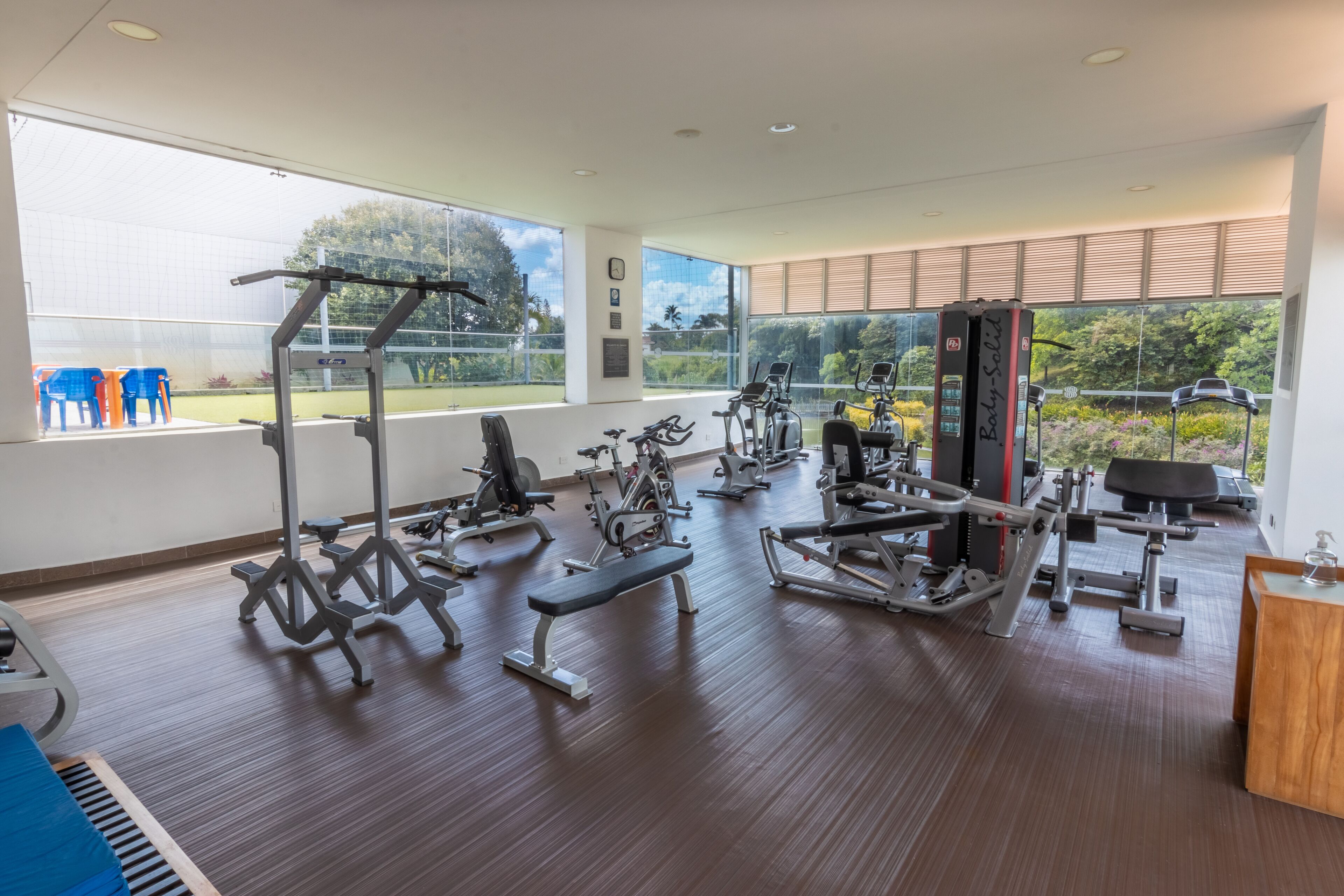Uma academia ampla com diversos equipamentos de exercício, amplas janelas com vista para uma quadra de tênis e piso de madeira.