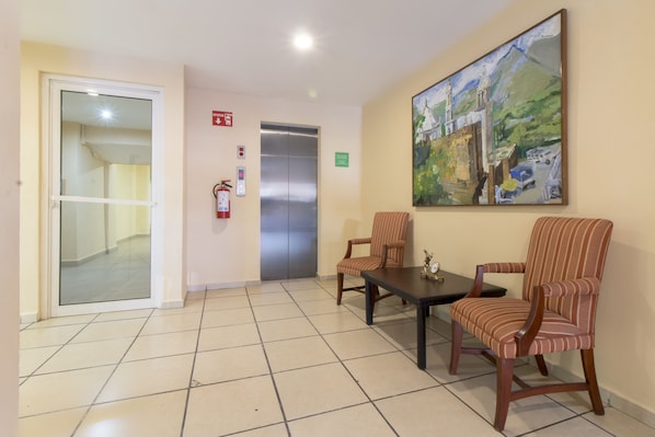 Um hall com chão de azulejos, duas cadeiras listradas, uma mesa pequena, um elevador e uma pintura na parede.