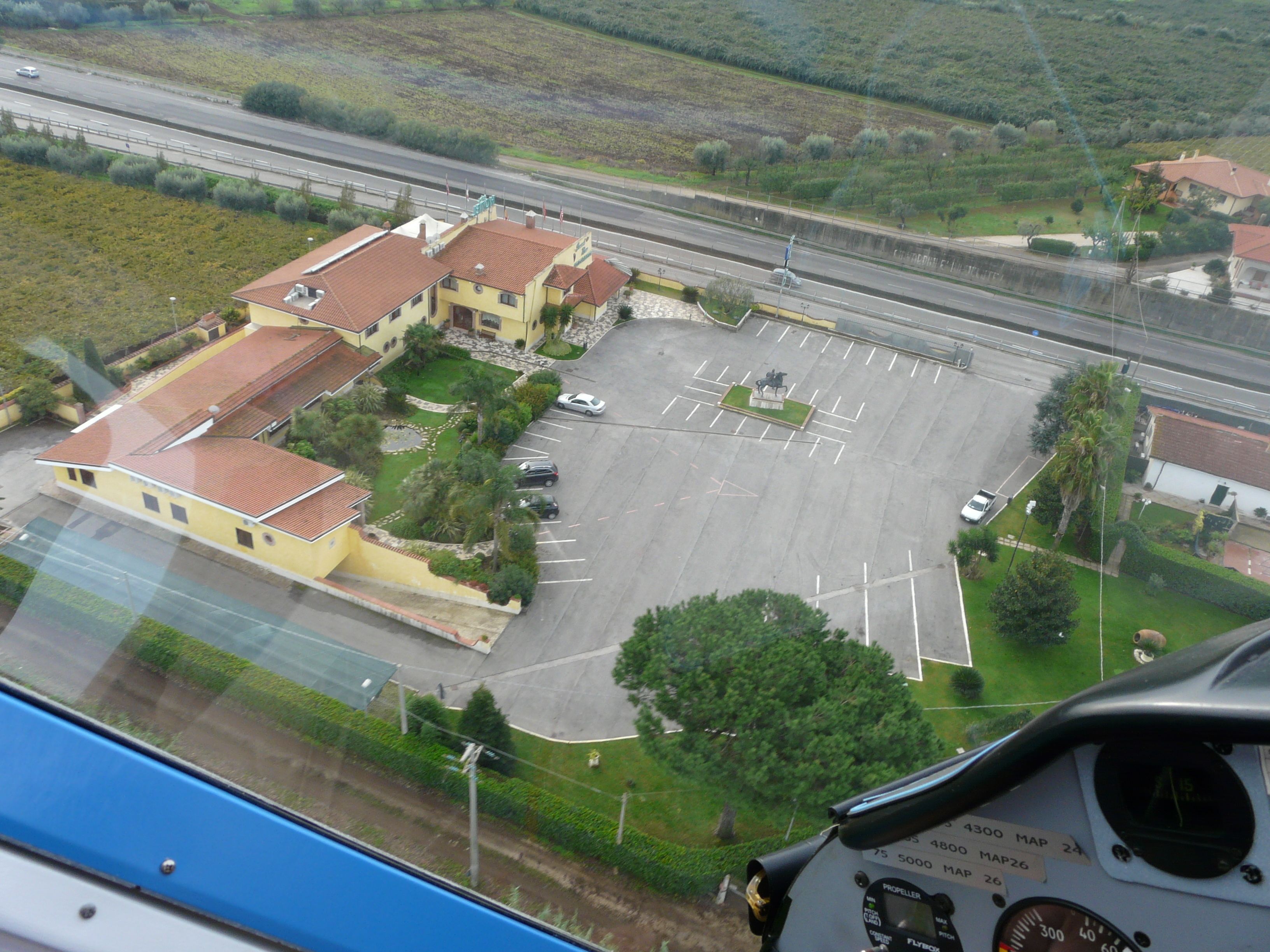 Vista aérea de um edifício com estacionamento, rodeado por vegetação e uma estrada.