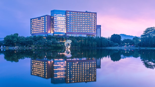 Um hotel moderno refletido na água, rodeado por árvores e um céu limpo.