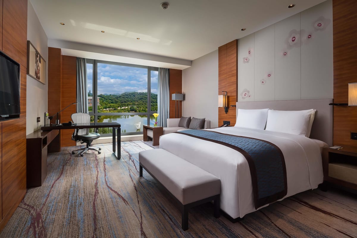 Quarto de hotel moderno com uma cama grande, uma escrivaninha, uma cadeira, um banco, uma TV e vista para um lago e colinas.