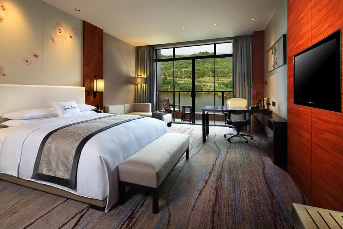Quarto de hotel com uma cama grande, uma mesa com cadeira, televisão e vista para um lago.