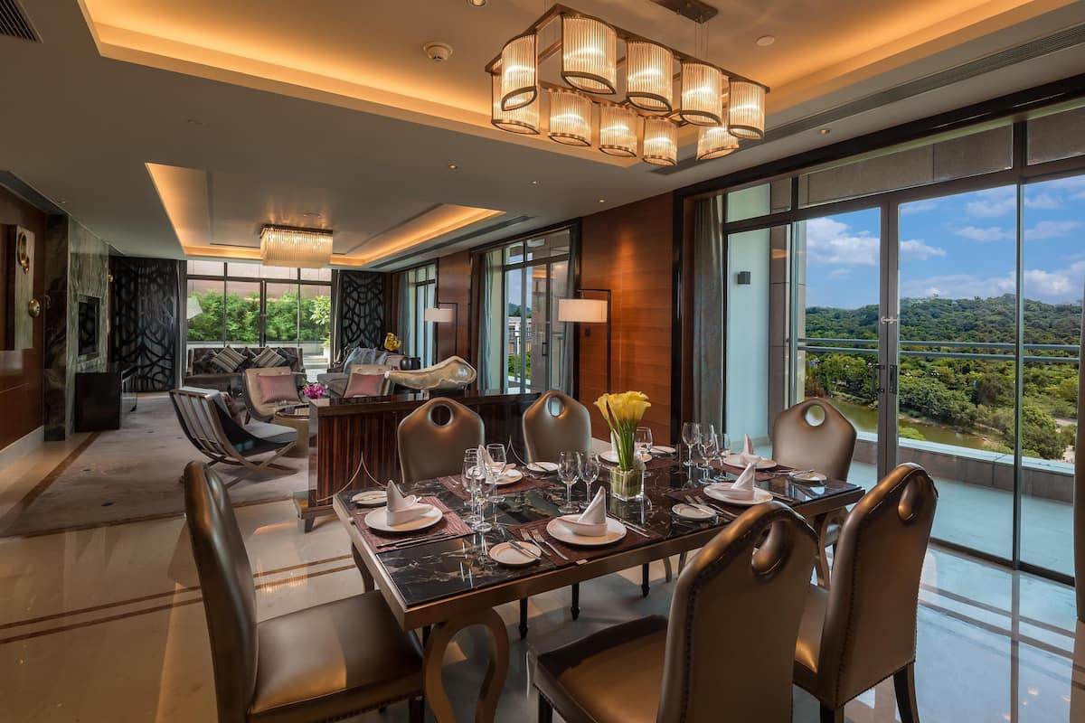 Uma sala de jantar com uma mesa grande posta para seis pessoas, rodeada por cadeiras, com vista para o verde através de grandes janelas.