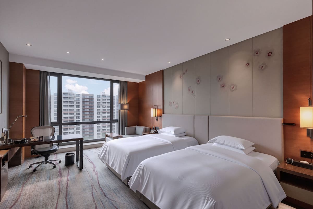 Quarto de hotel com duas camas, uma escrivaninha, uma cadeira e uma janela ampla com vista para a cidade.