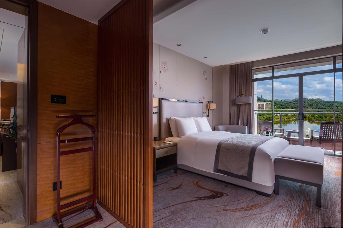 Quarto de hotel com cama, mesa de cabeceira, abajur e vista para uma varanda com mobiliário.
