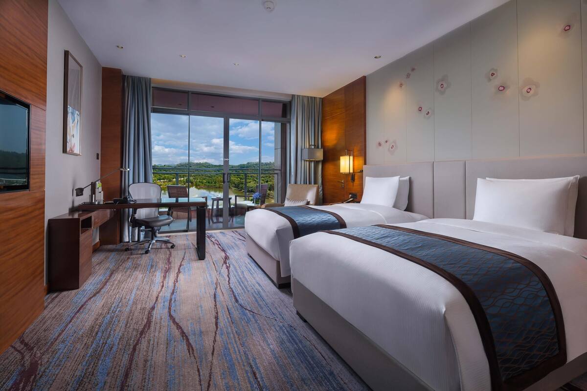 Quarto de hotel com duas camas, uma escrivaninha, uma cadeira, televisão e uma janela grande com vista.