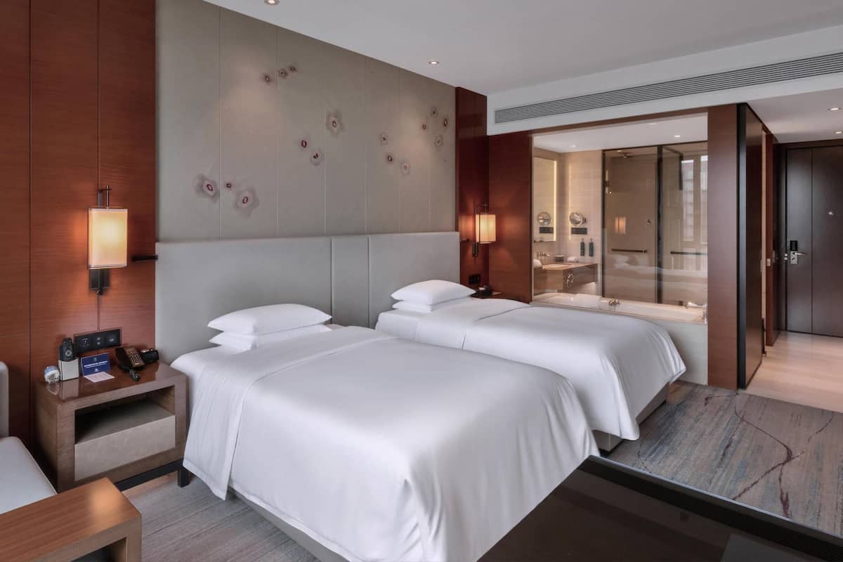 Quarto de hotel com duas camas, uma escrivaninha, uma cadeira, banheiro com chuveiro e porta que dá para outro quarto.