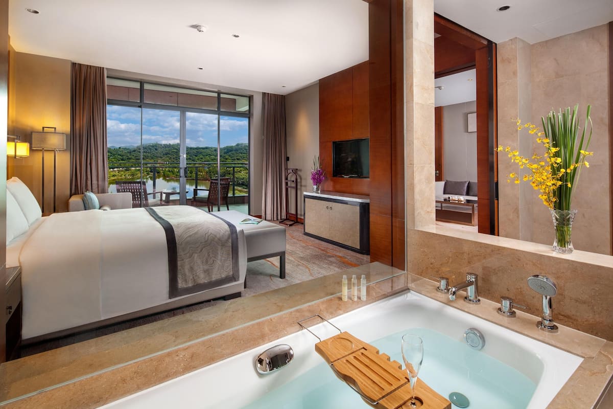 Quarto de hotel moderno com uma cama grande, um espaço de estar, banheiro com banheira e vista para o exterior.