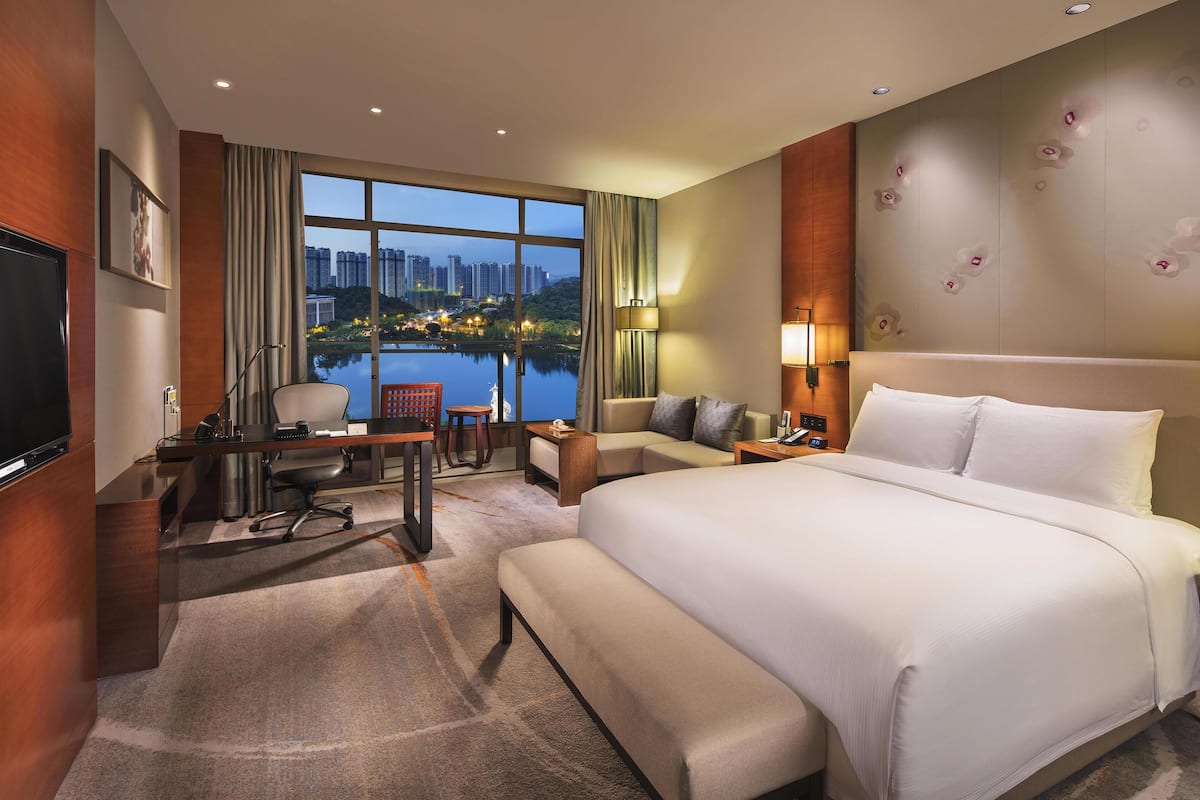 Quarto de hotel com uma cama grande, uma mesa com cadeira, uma televisão e vista para a paisagem urbana através de amplas janelas.