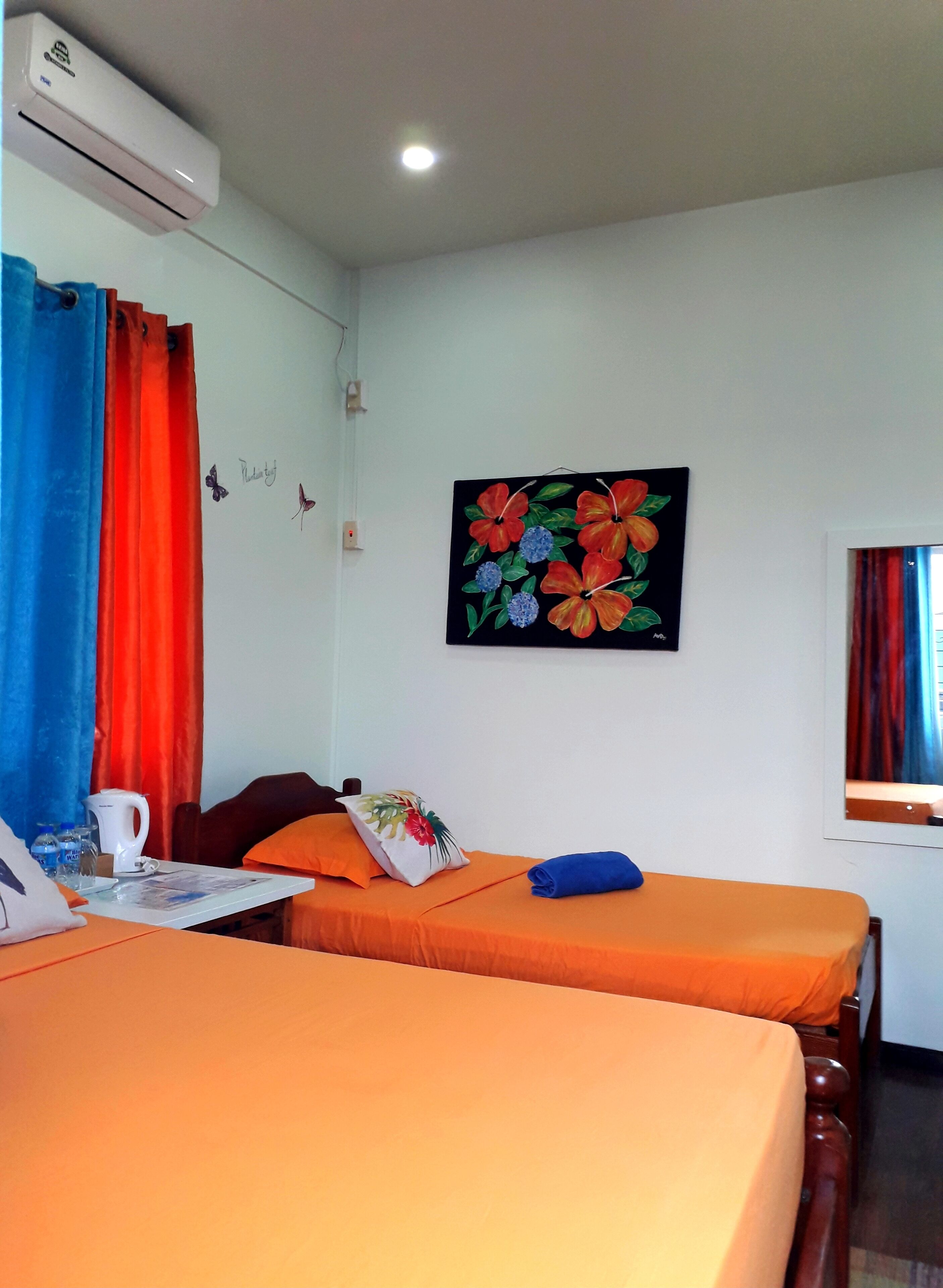 Quarto de hotel com duas camas, unidade de ar condicionado, pintura de flores e janela com cortinas.