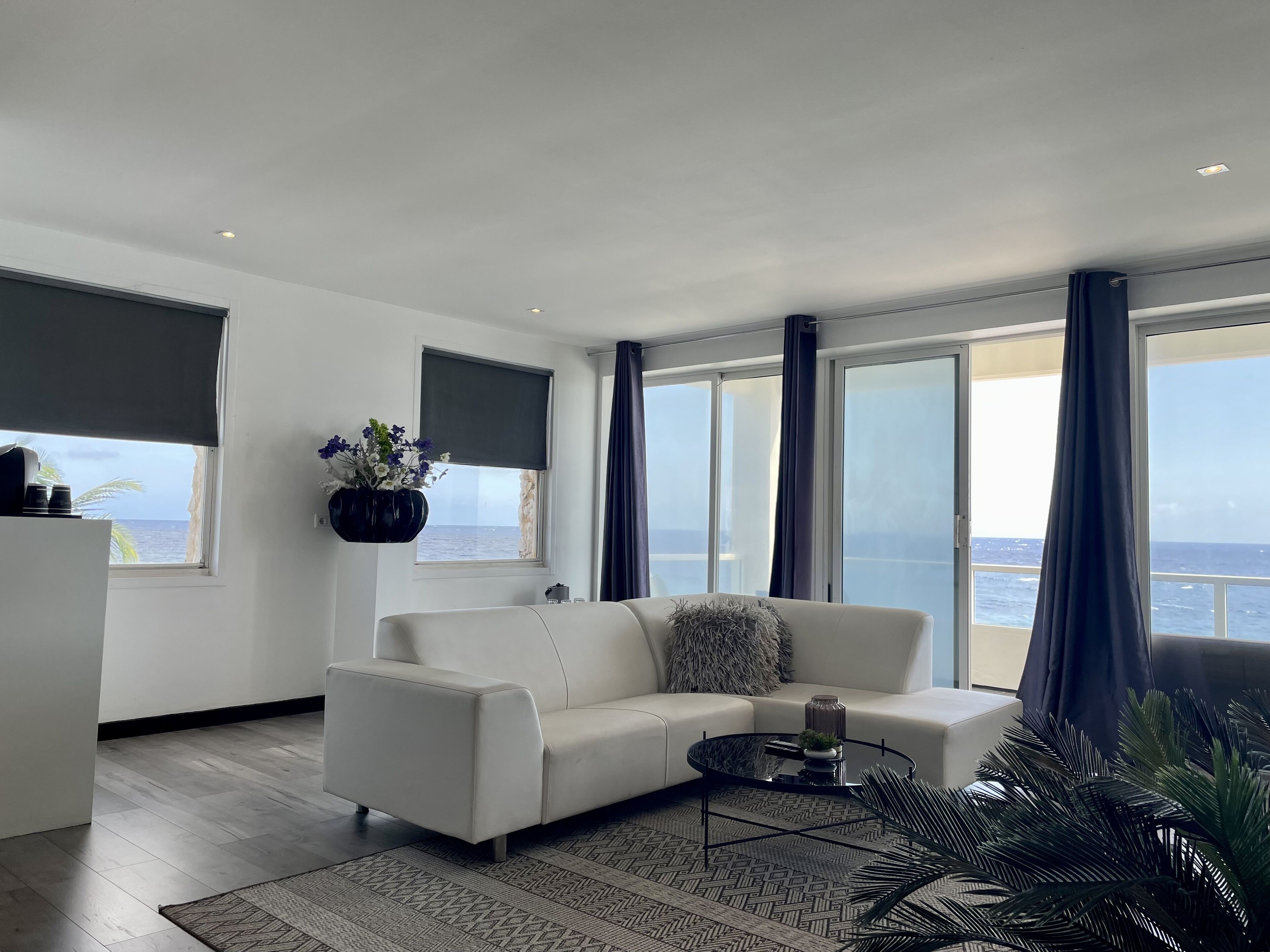 Uma sala de estar moderna com um sofá branco, uma mesa de centro redonda e vista para o oceano por grandes janelas.
