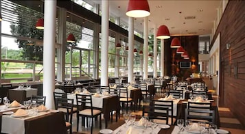 Um restaurante espaçoso com pé direito alto, janelas amplas e iluminação pendente vermelha. Mesas organizadas com toalhas brancas e preparadas para refeições.