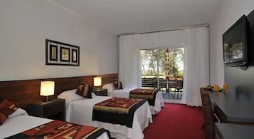 Quarto de hotel com duas camas, televisão, varanda com mesa e cadeiras, e janela com cortinas.