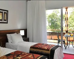 Quarto de hotel com duas camas, uma mesa de cabeceira com abajur, uma janela grande com cortinas e uma varanda com mesa e cadeiras.
