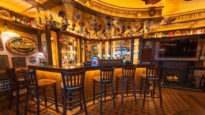 Um bar com chão xadrez, balcão de madeira e banquetas. Vários garrafas e uma tela de televisão na parede.