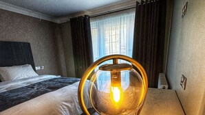 Quarto de hotel com cama, mesa de cabeceira com abajur e janela com cortinas.