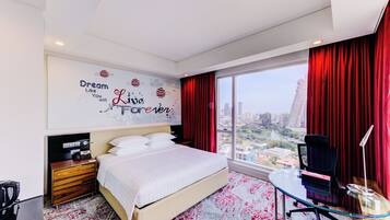 Quarto de hotel moderno com uma cama grande, uma escrivaninha com cadeira, vista para a cidade e arte motivacional na parede.