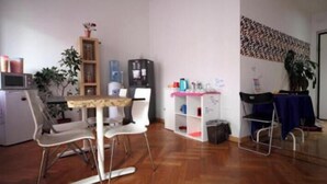 Quarto com piso de madeira, paredes brancas, uma escrivaninha branca com cadeira, um bebedouro, uma geladeira e uma mesa azul com cadeiras.