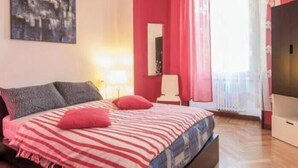 Quarto com cama, mesa de cabeceira, luminária, espelho, televisão e janela com cortinas.