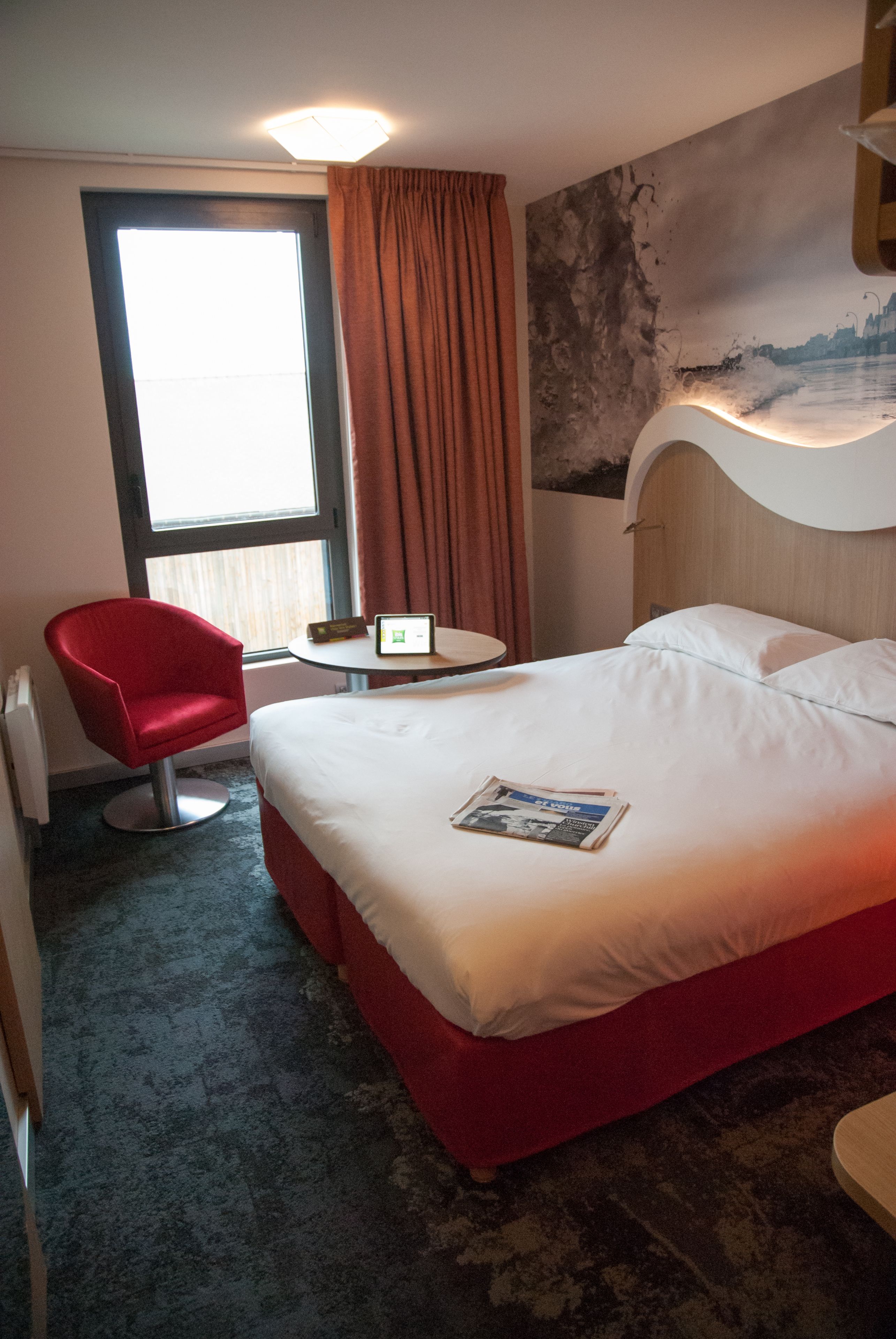Quarto de hotel com uma cama grande, uma cadeira vermelha, uma mesinha com abajur e um quadro na parede.