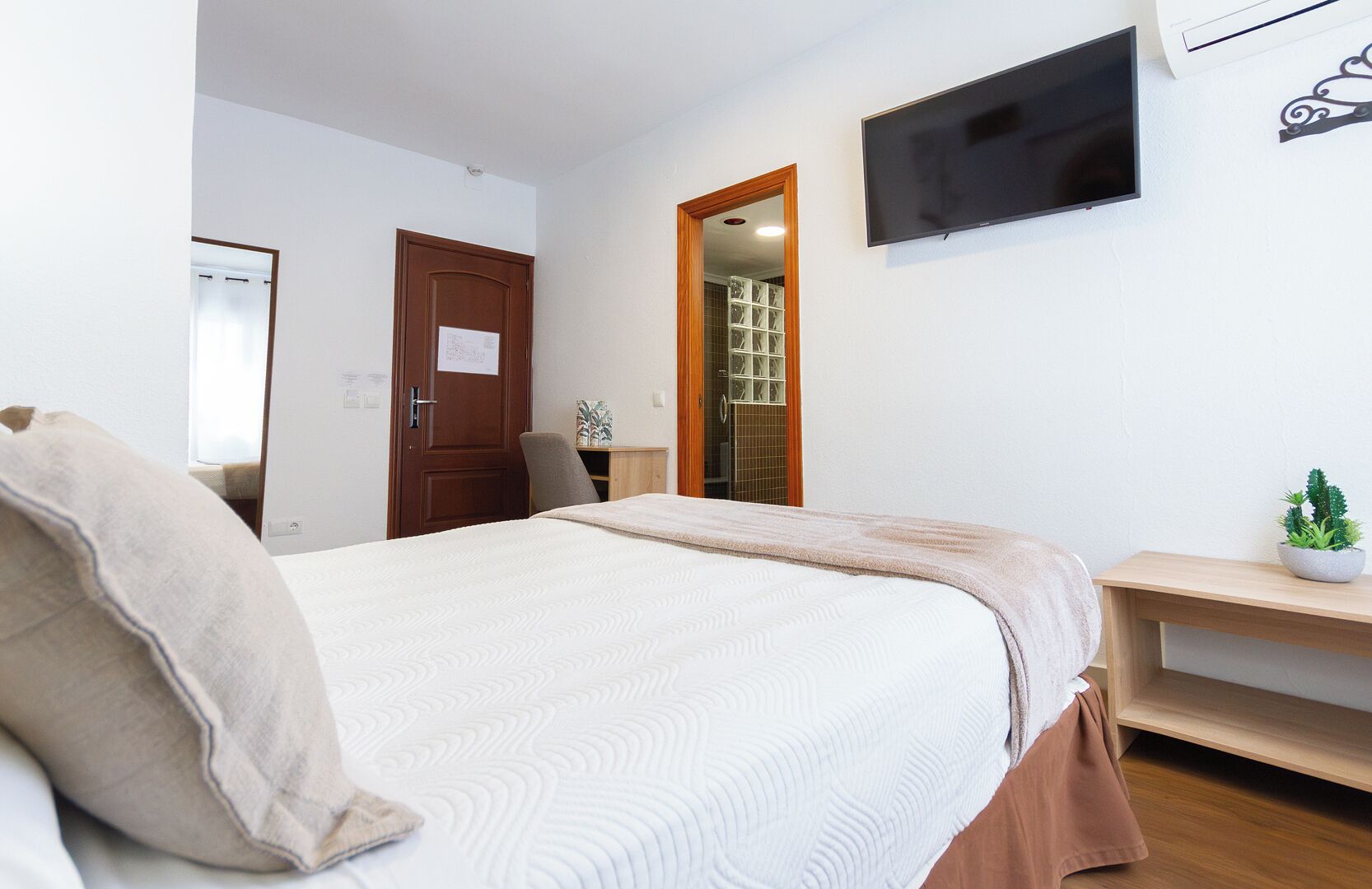Quarto de hotel com uma cama, uma televisão fixada na parede, uma porta de madeira e uma pequena planta sobre uma mesinha de cabeceira.