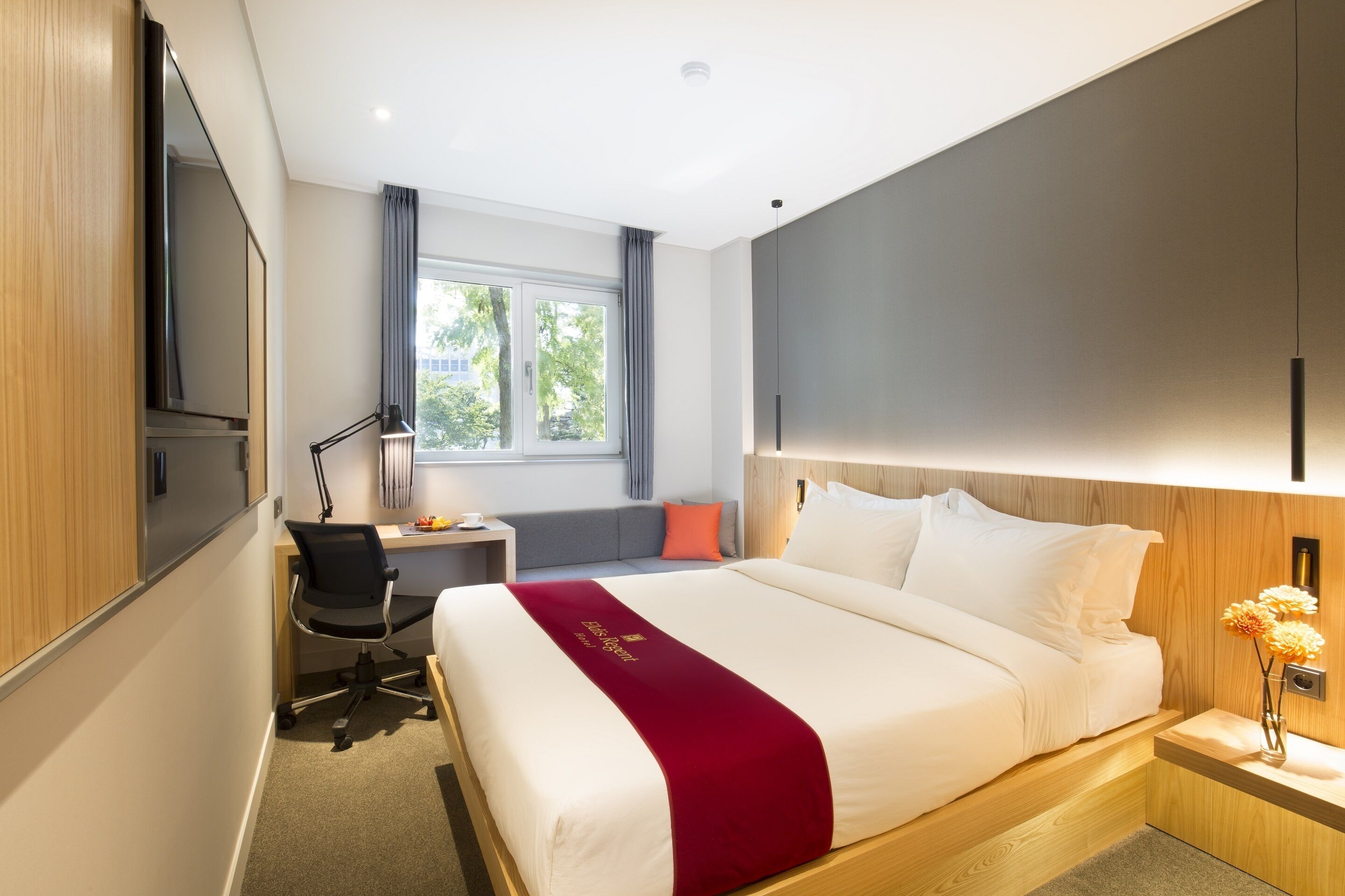 Quarto de hotel moderno com uma cama grande, uma escrivaninha com cadeira, uma mesinha, um espelho e uma janela com cortinas.